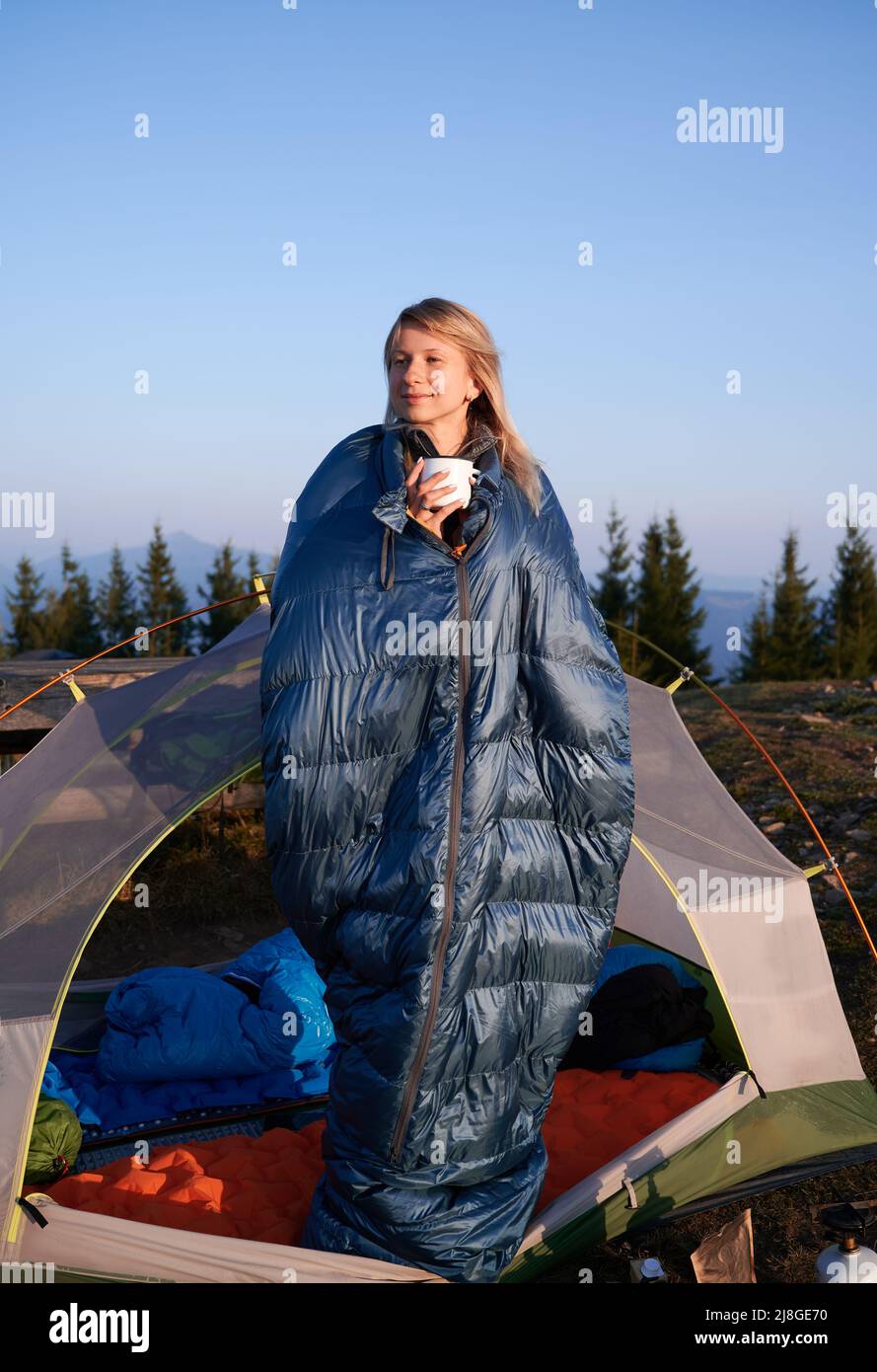 Young woman wrapped in sleeping bag standing against blue sky, trees ...