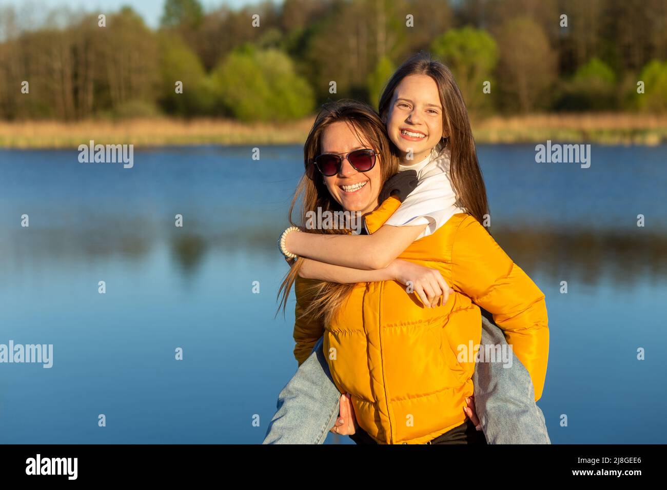 Playful mother giving daughter piggy back ride at spring lake shore ...