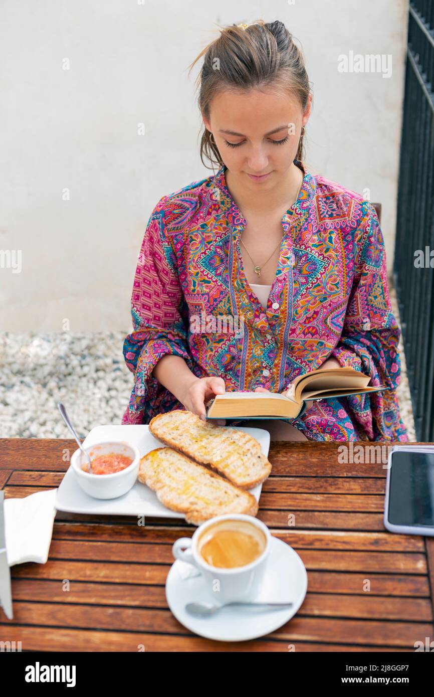 woman having toast for breakfast on a bar terrace and reading a book ...