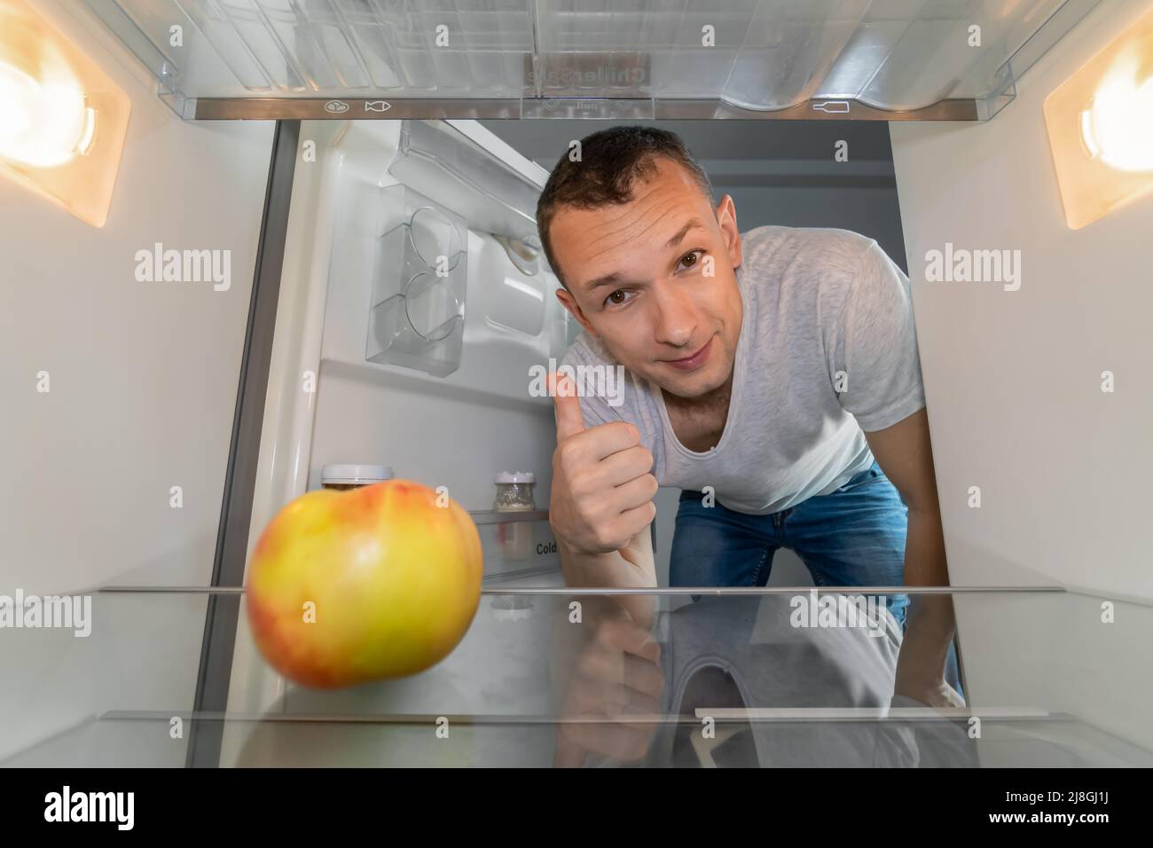 Smiling man looks inside the refrigerator with one apple, shows a ...