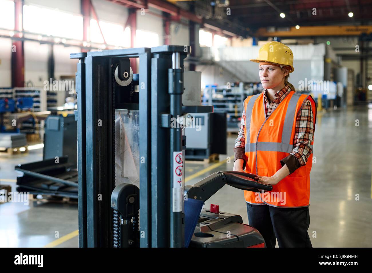 Young specialist of large modern factory pushing forklift along wide ...