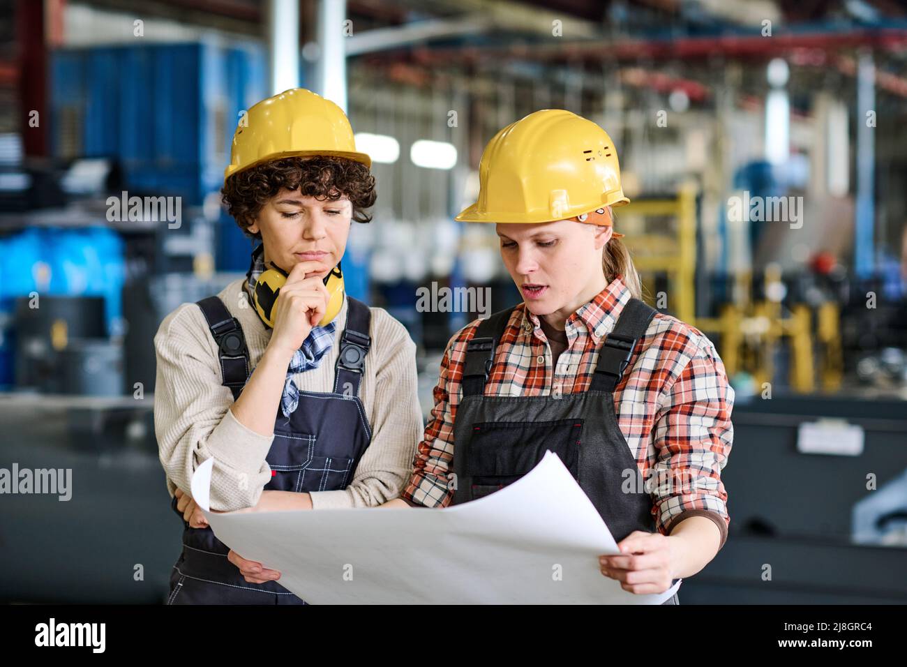 Pensive female engineer looking at sketch on blueprint held by colleague explaining its details ...