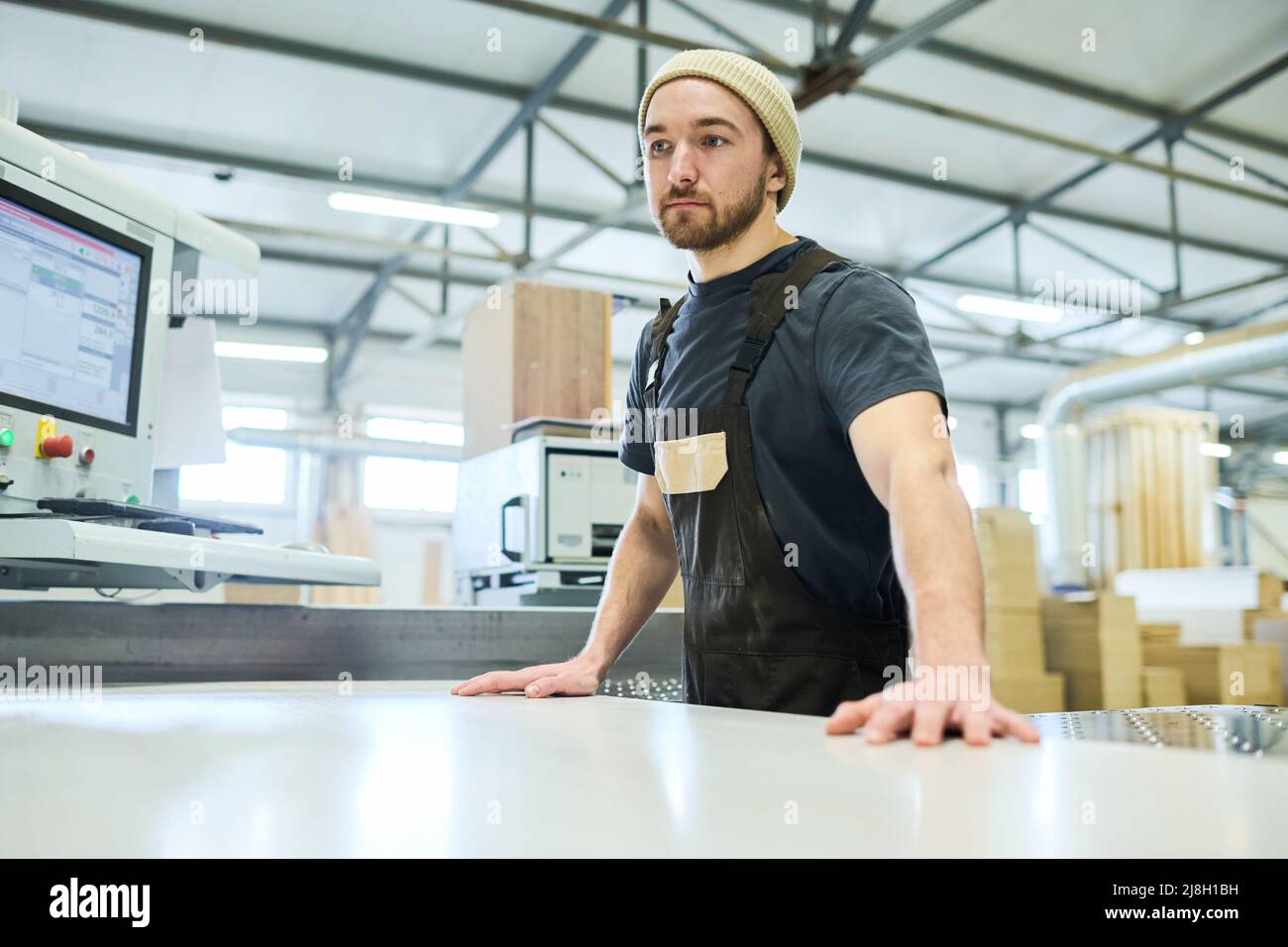 Young carpenter in overall standing at machine equipment and doing wood ...