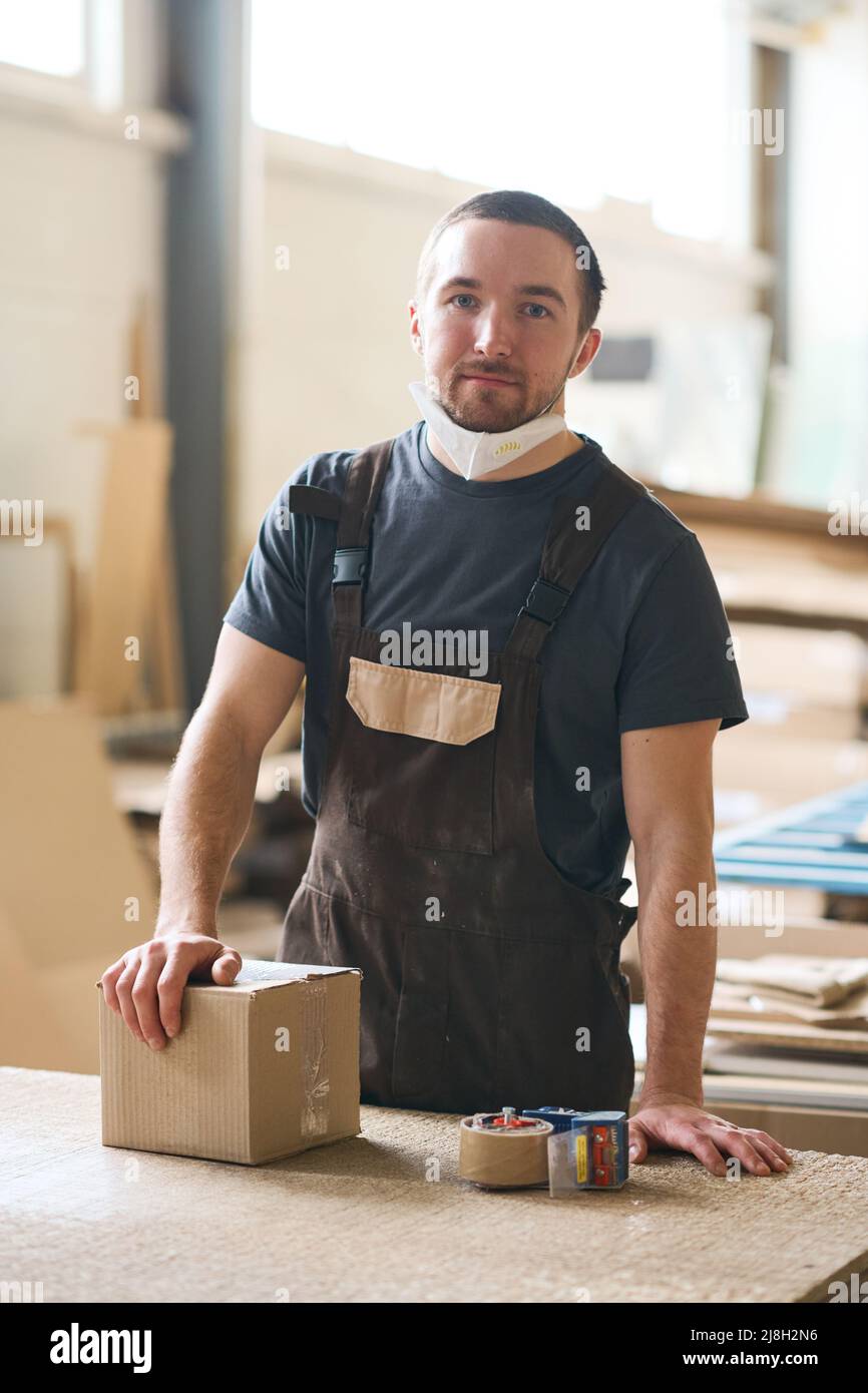 Portrait of young worker in overall looking at camera while packing box ...