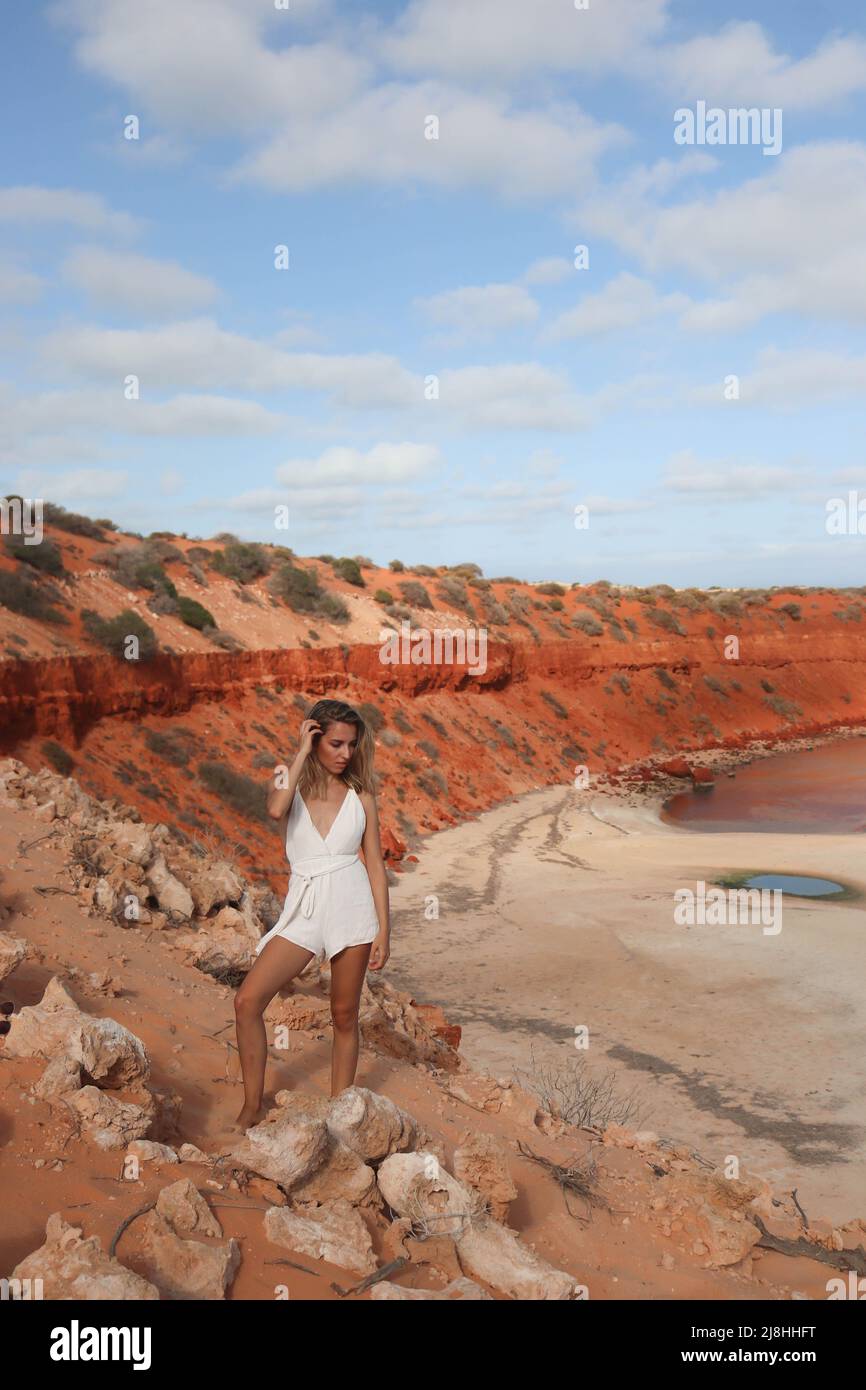 Pretty young blonde woman in white dress on cliff with red sandy beach ...