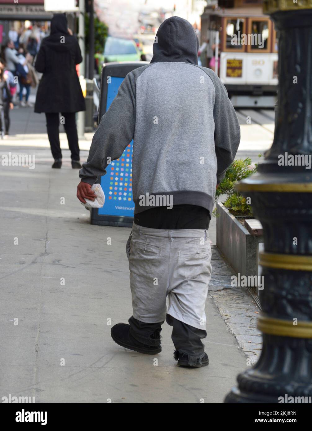 A homeless man wearing two pairs of pants walks along a sidewalk in ...