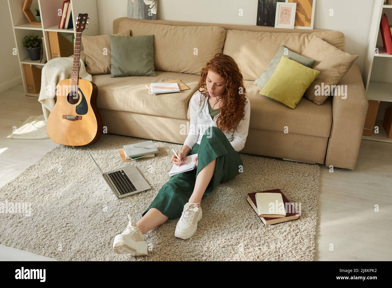 Above view of curly-haired girl sitting on carpet and making notes ...
