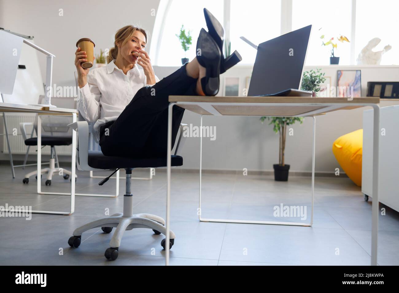 Business woman with a cup of coffee put her legs on the desk and eats ...