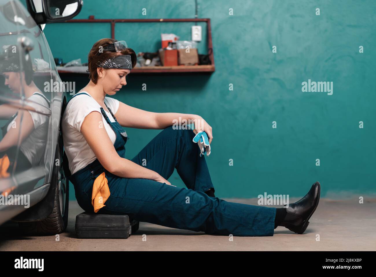 A tired sad caucasian woman mechanic is sitting leaning on a car in an ...