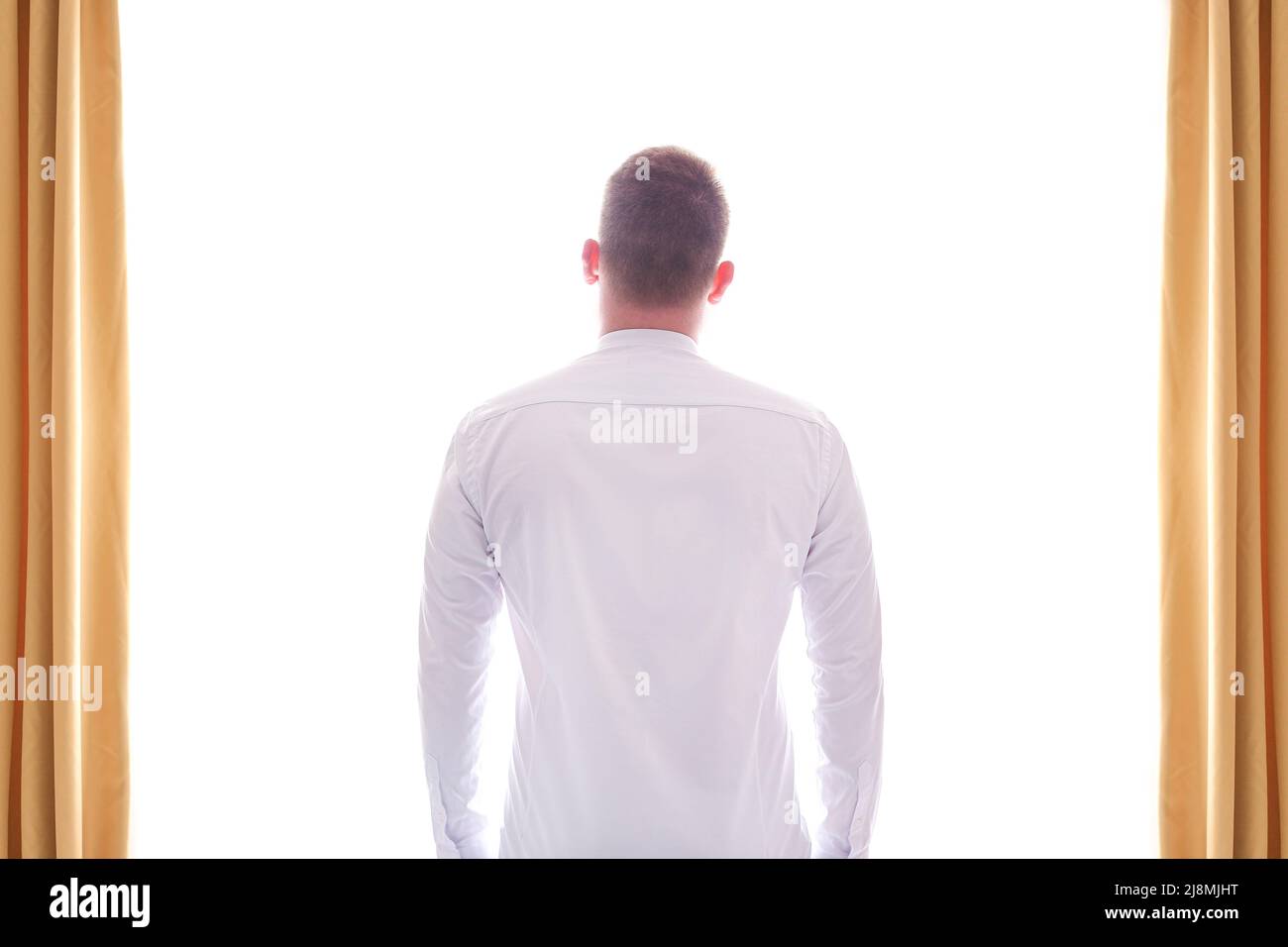 A man in a white shirt stands with his back looking out the window with ...
