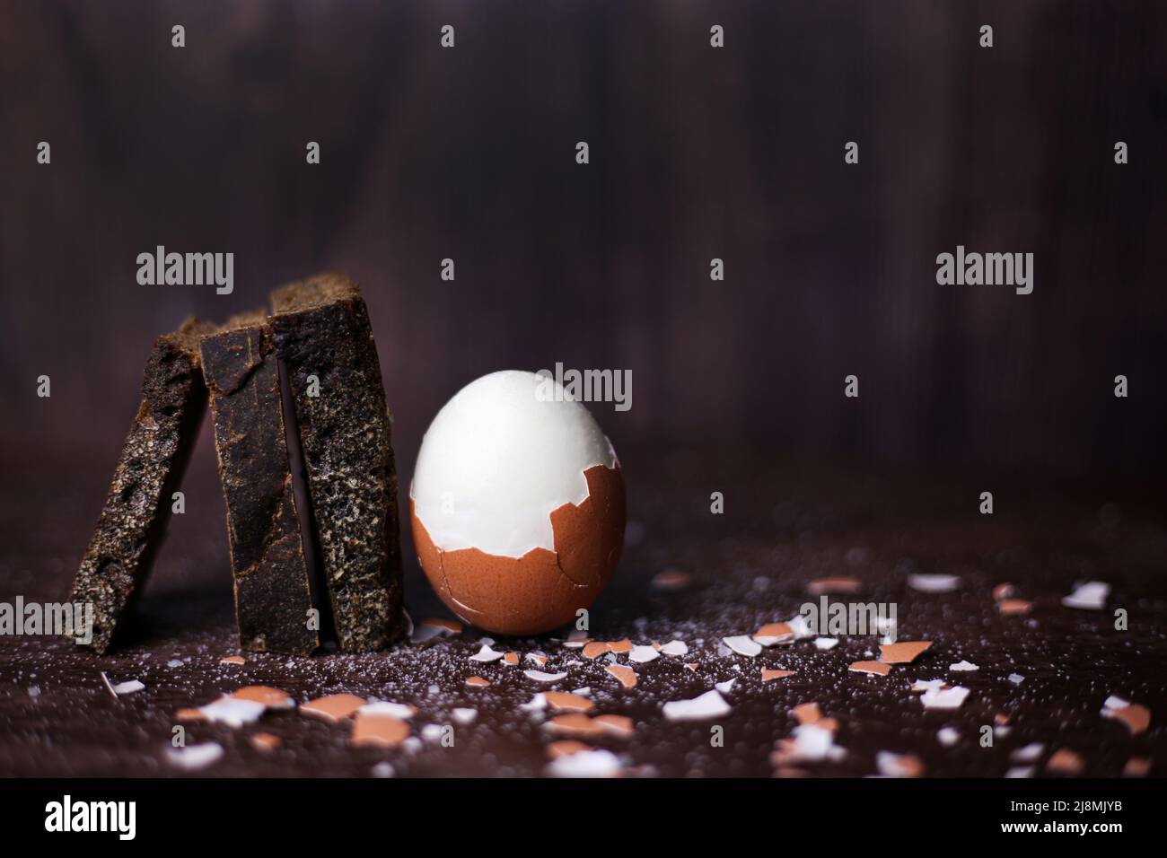 Brown boiled half-peeled egg and pieces of black bread on a dark wooden ...