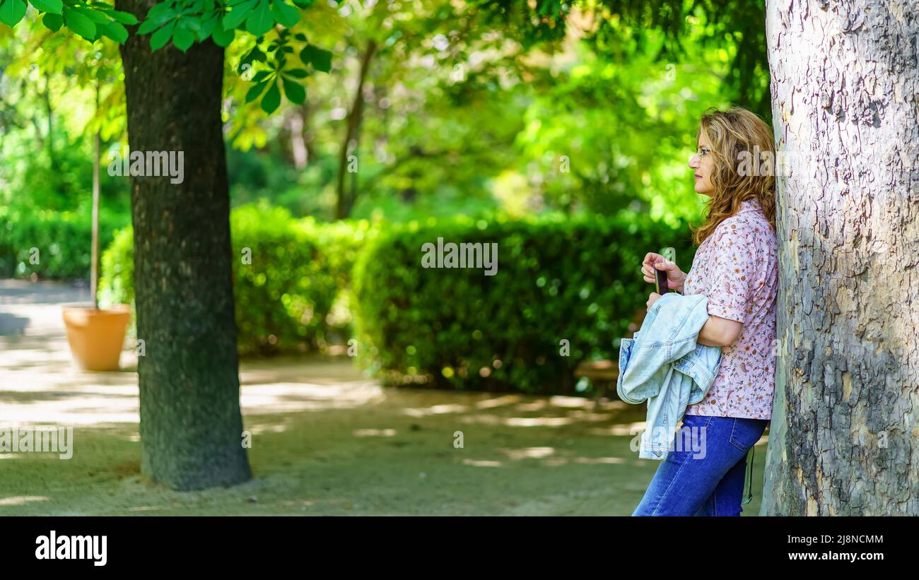 Senior woman leaning on the trunk of a huge tree in the light of the ...