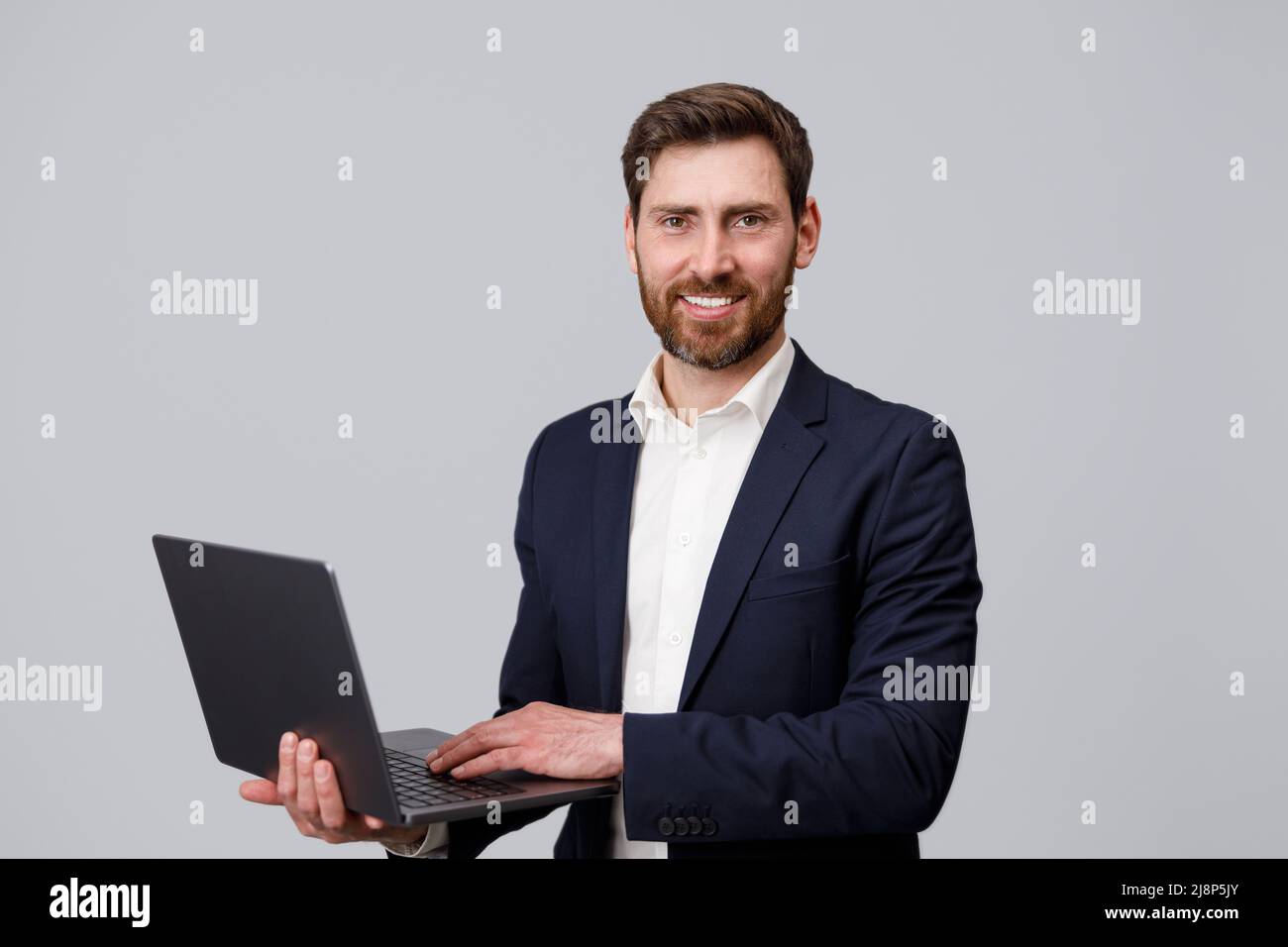 Smiling male business coach in suit holding laptop over grey studio ...