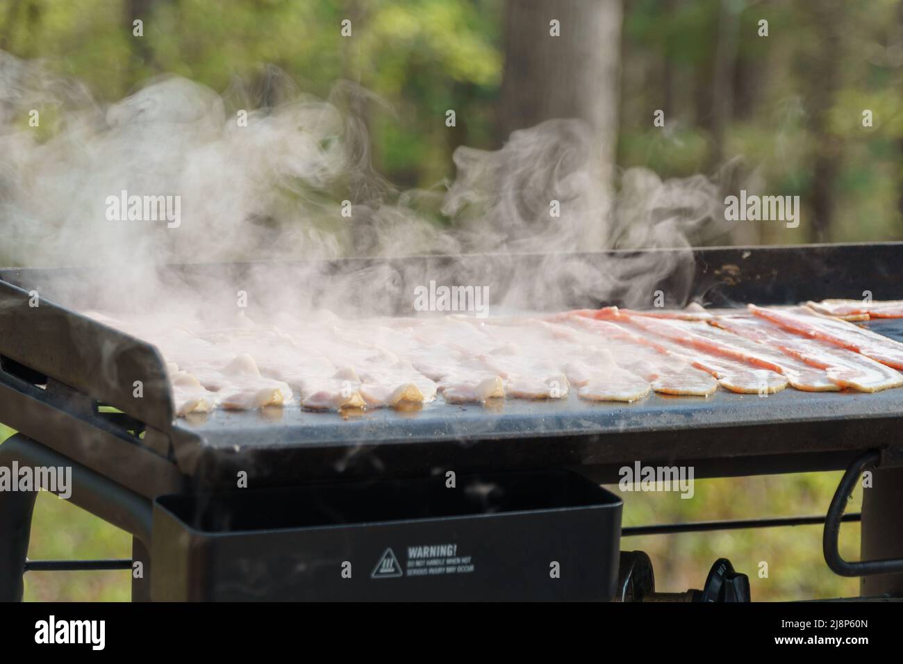 Bacon cooking on an black outdoor camping grill with steam rising and ...