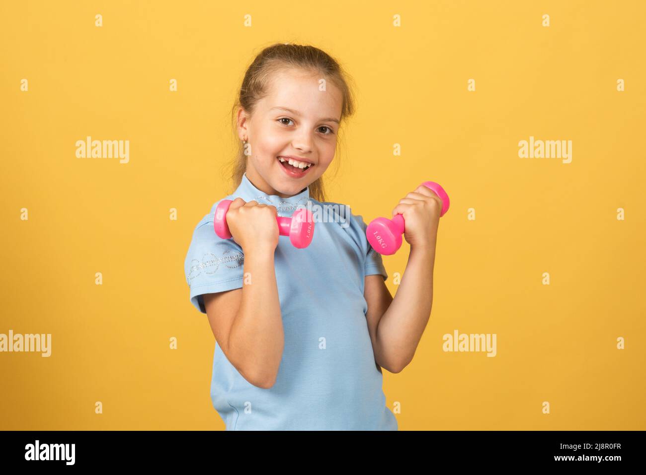Cute little girl doing exercises with dumbbells in yellow background ...