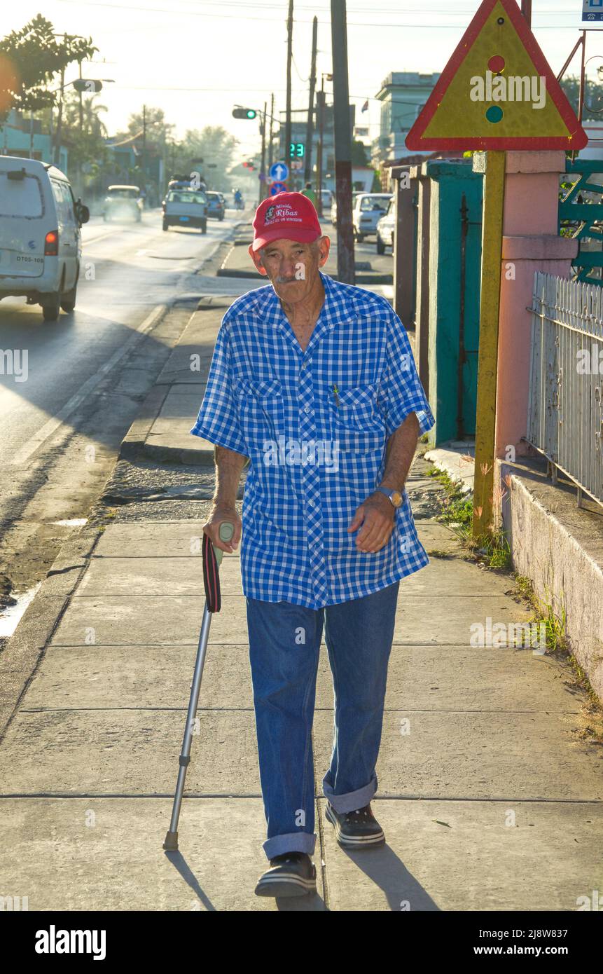 Senior Cuban man with a cane walking in the sidewalk. He wears a cap ...
