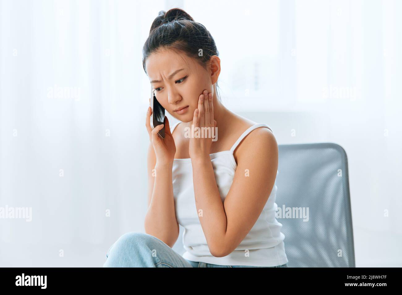 Wary irritated angry tanned lovely young Asian woman listen bad news at call using phone at home ...