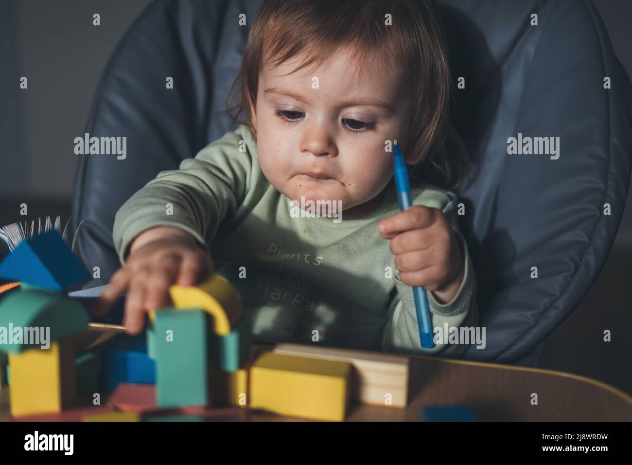 Baby girl playing with multicolored building blocks and holding a pen ...