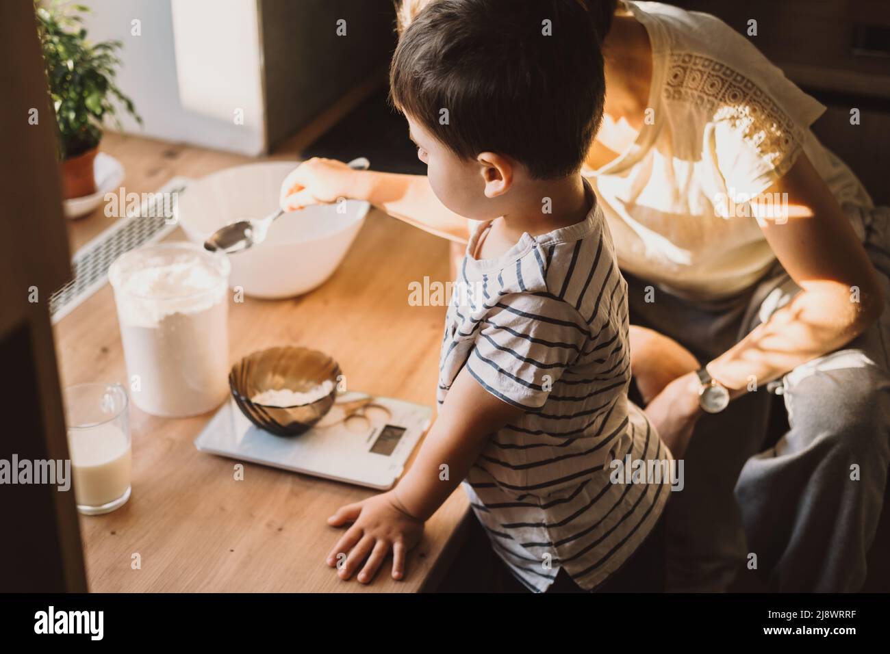 Boy helping his mother make cake dough by adding flour with a spoon ...