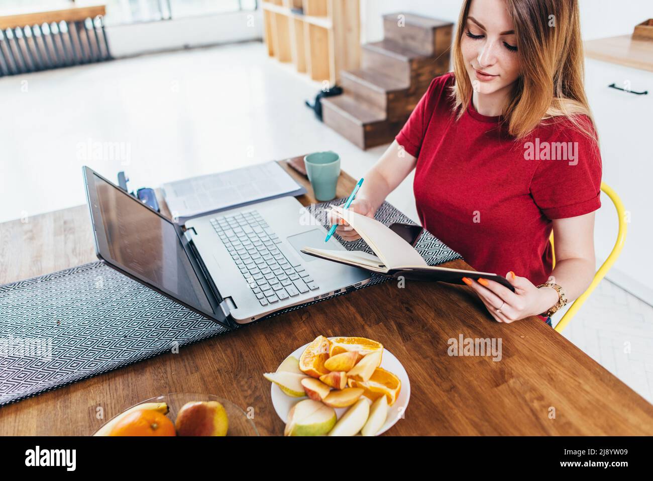 Woman writing and taking notes, using her laptop in kitchen. Working at ...