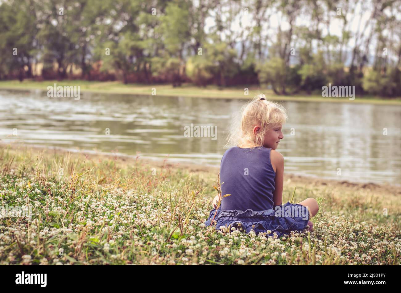 little girl sitting back view in green meadow and looking to the pond ...