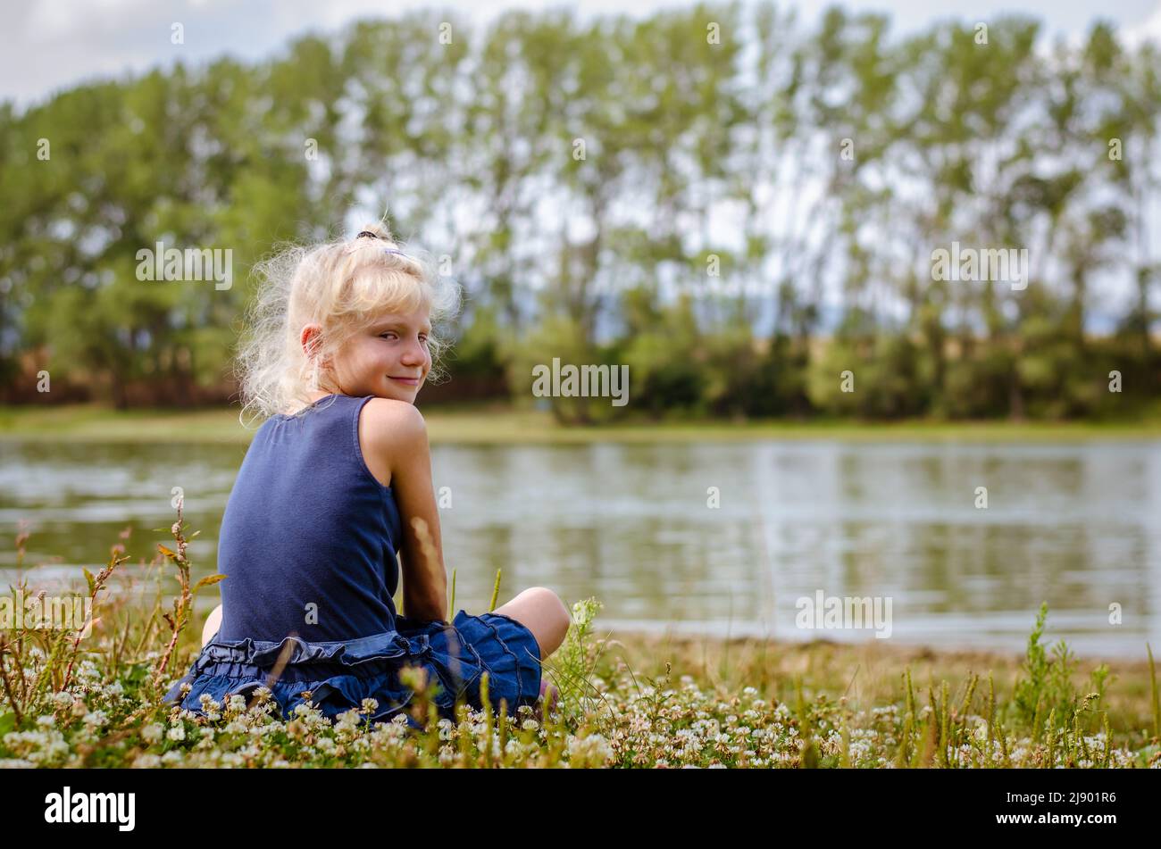 little girl sitting back view in green meadow and looking to the pond ...