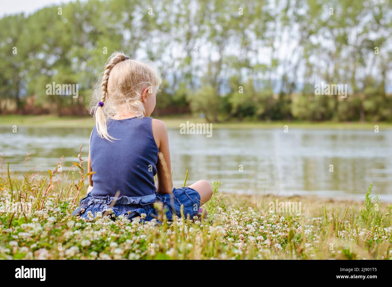 little girl sitting back view in green meadow and looking to the pond ...