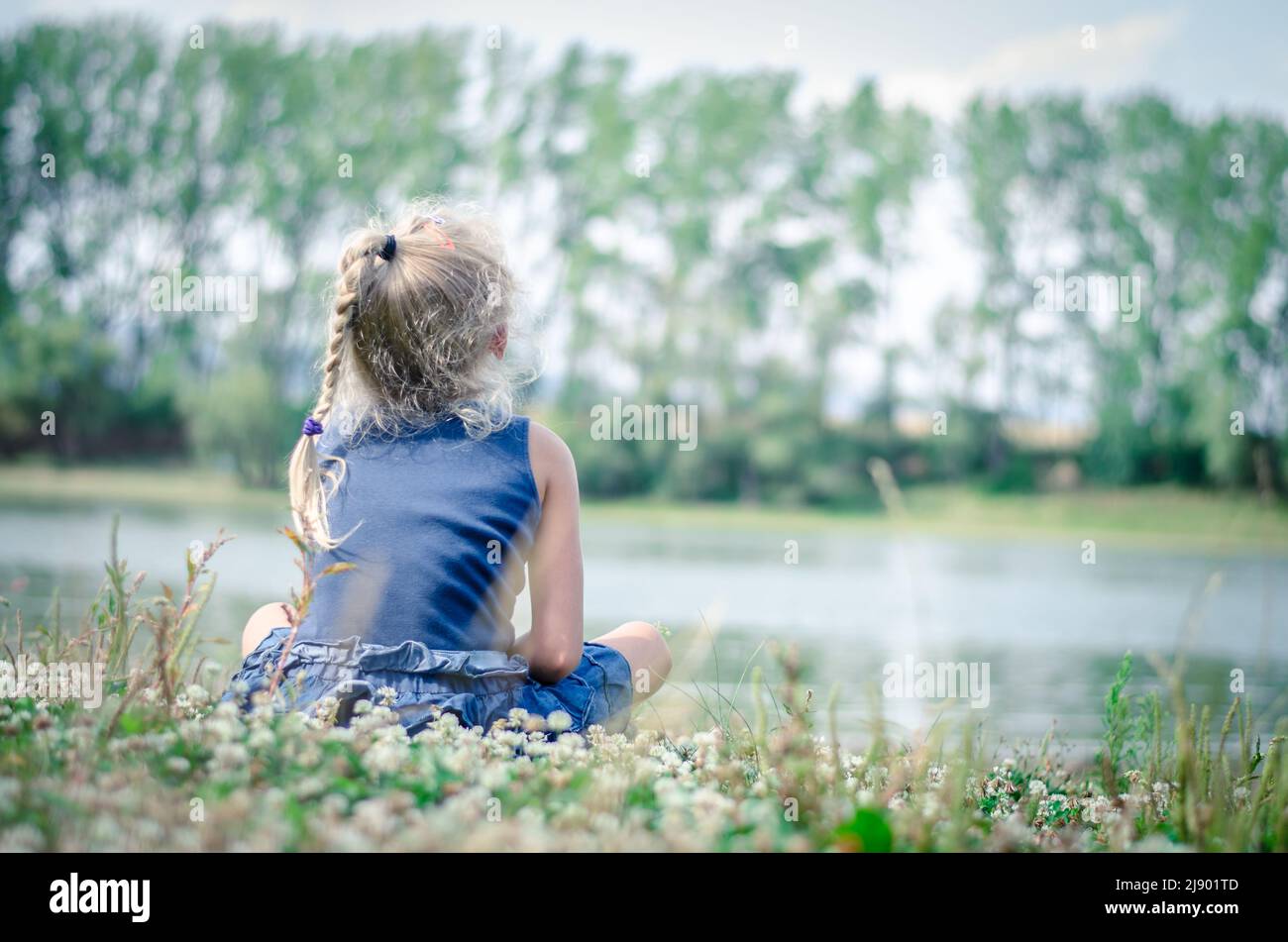 little girl sitting back view in green meadow and looking to the pond ...