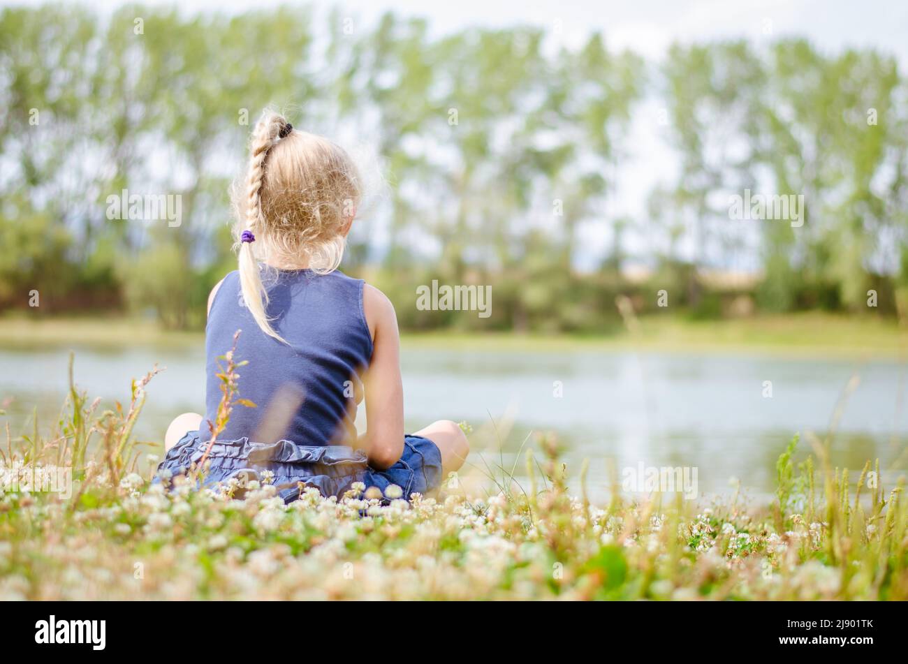 little girl sitting back view in green meadow and looking to the pond ...