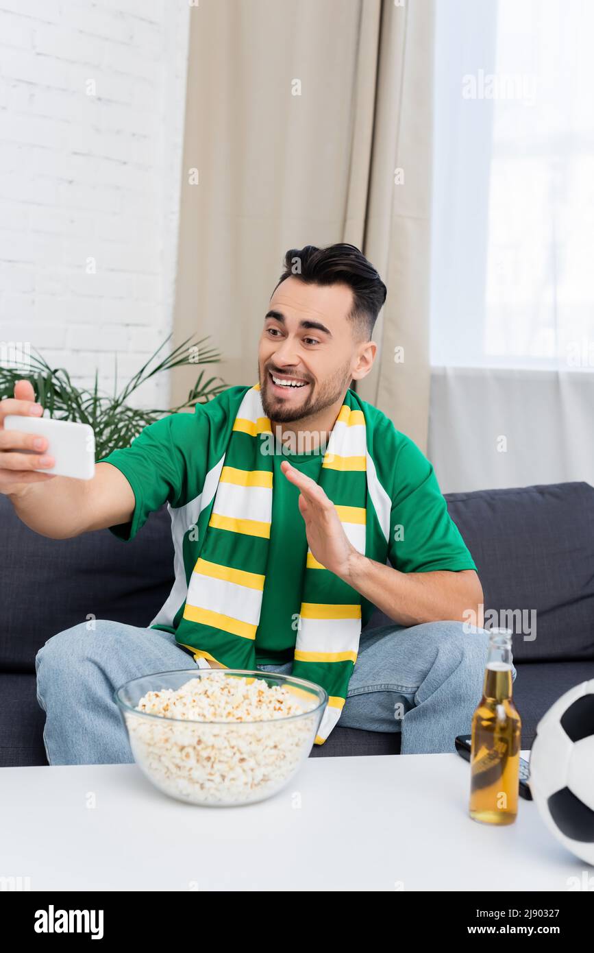 cheerful sports fan waving hand during video call near popcorn and beer ...