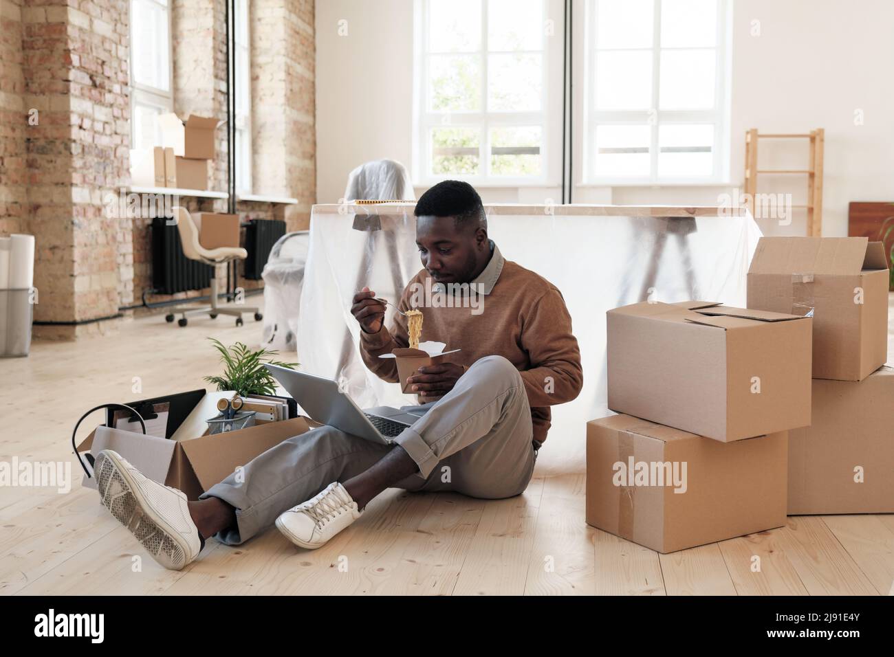 Young Afro-American man sitting among cardboard boxes and using laptop ...