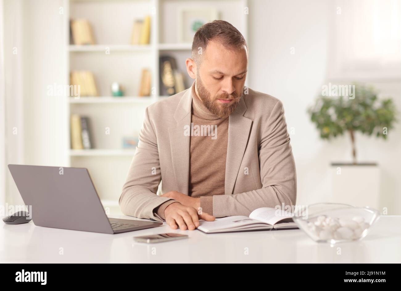 Man sitting at a desk with a laptop computer and reading a book at home ...