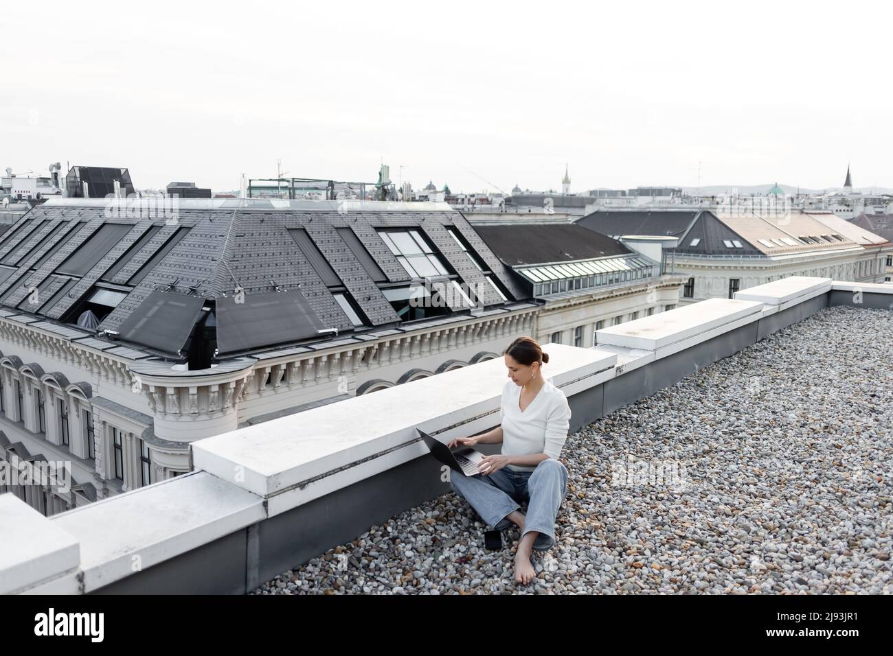 full length of barefoot woman in jeans working with laptop on rooftop ...