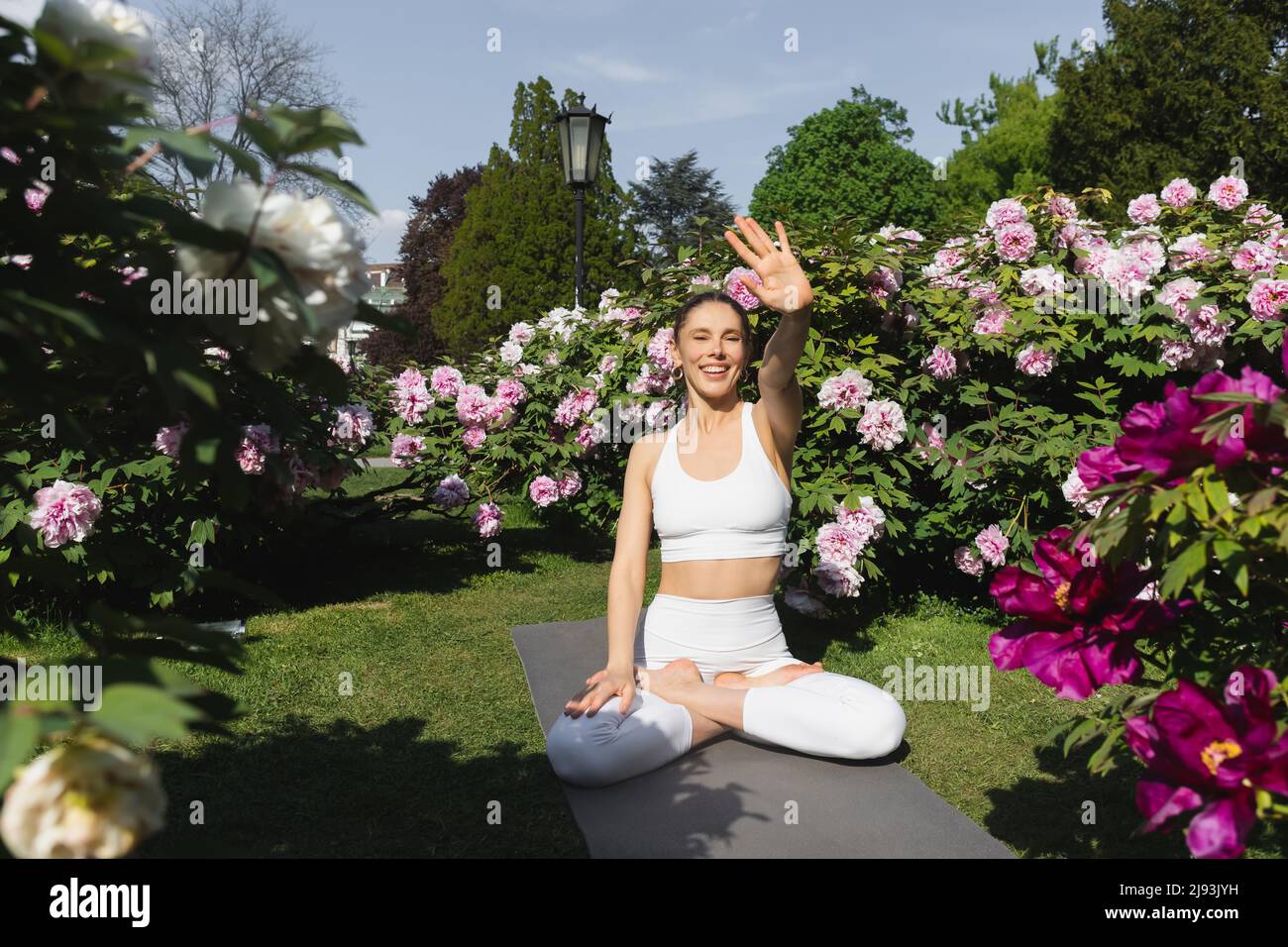happy woman sitting in lotus pose and waving hand near blossoming ...