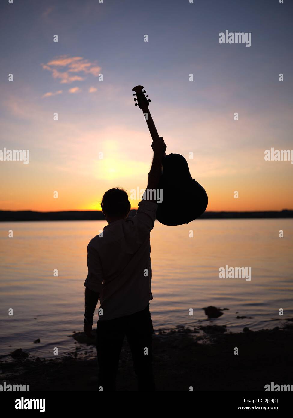 male guitarist with his guitar in the air at dusk in front of a lake ...
