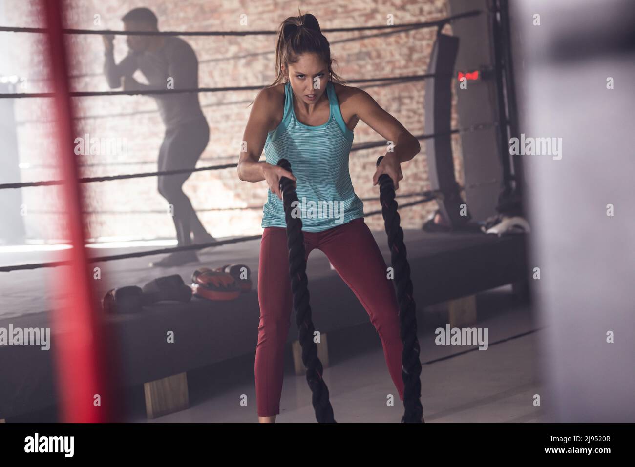 Dedicated caucasian young female boxer exercising with battle ropes in ...
