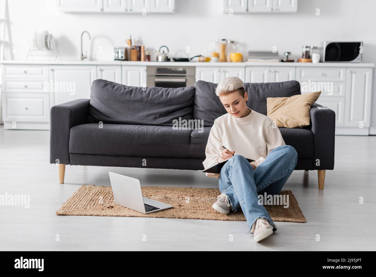 full length of woman in jeans writing in notebook near laptop on floor ...