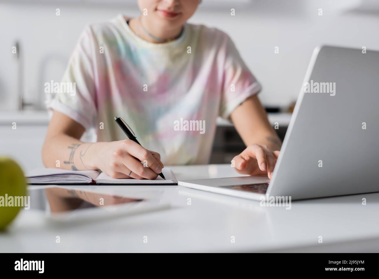 partial view of blurred woman writing in notebook near laptop and ...