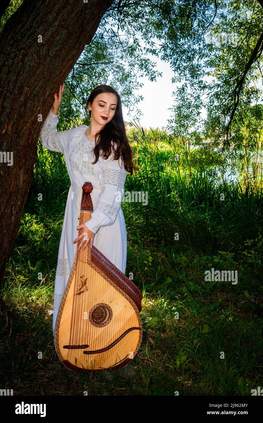 Ukrainian woman in a white dress stands under a tree by the river ...