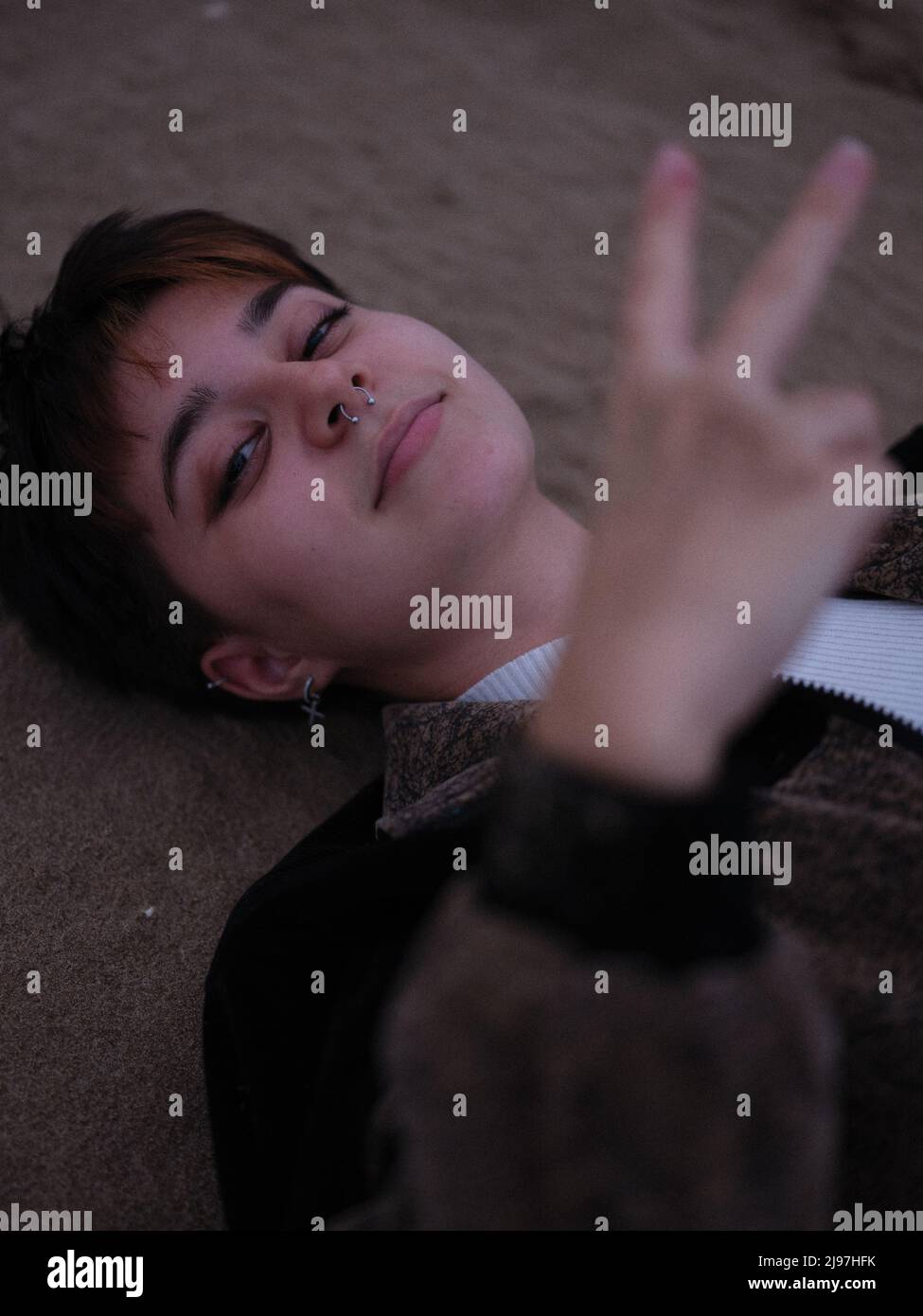 non-binary woman lying with her head on the sand, making a v-sign with ...