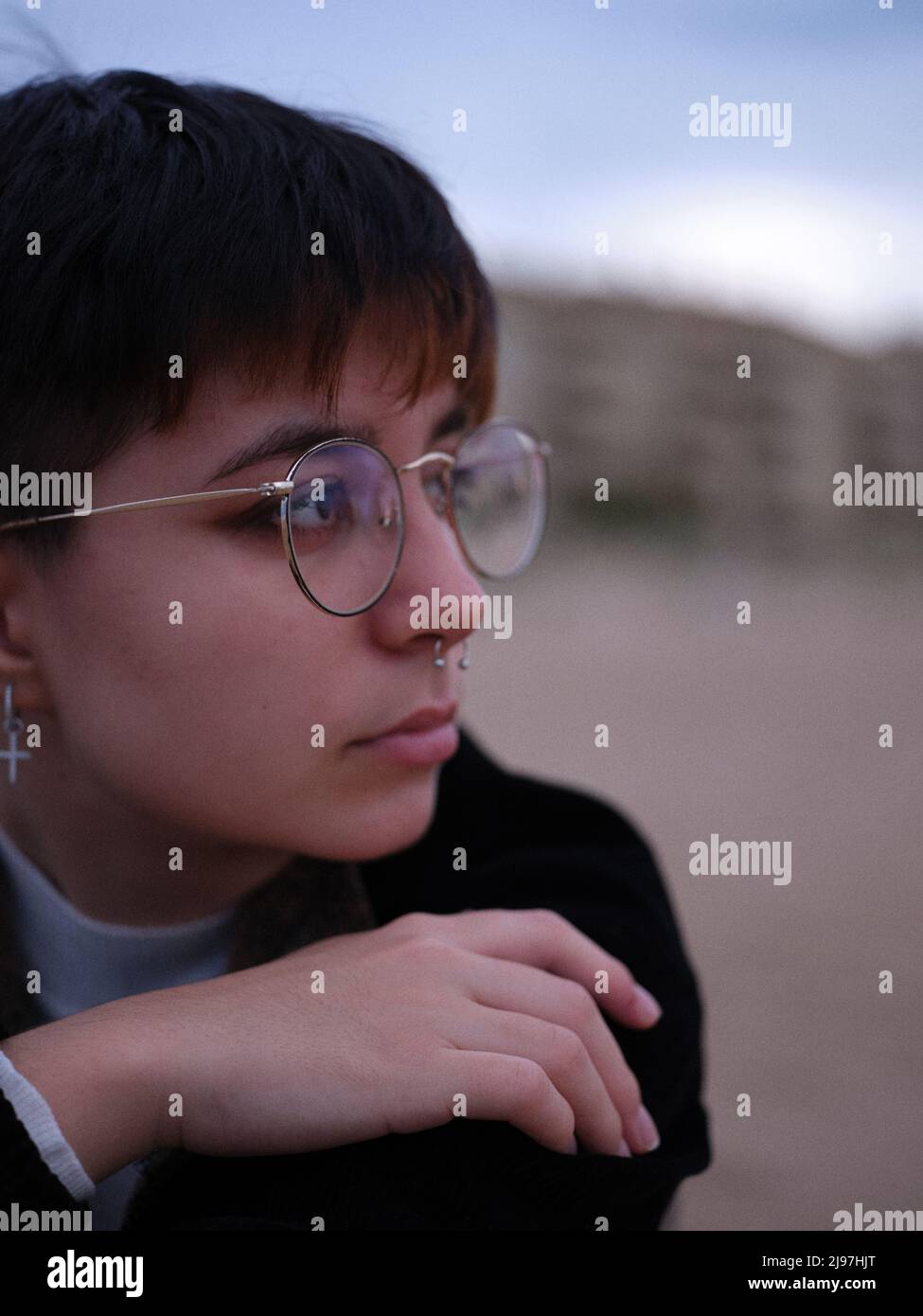 trans woman with glasses, piercings and short hair looking to the side ...