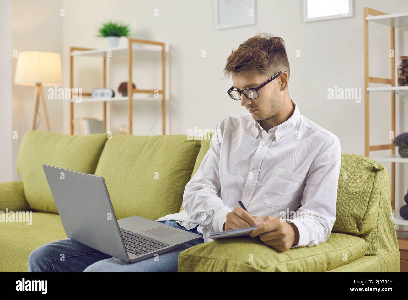 Young man sitting on sofa with laptop, having online class and taking ...