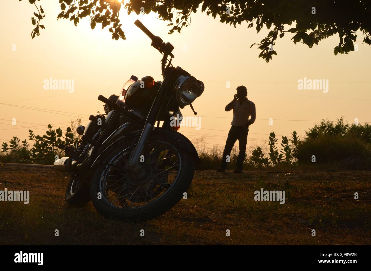 Silhouetted man chatting on a mobile phone behind his motorcycle in the ...