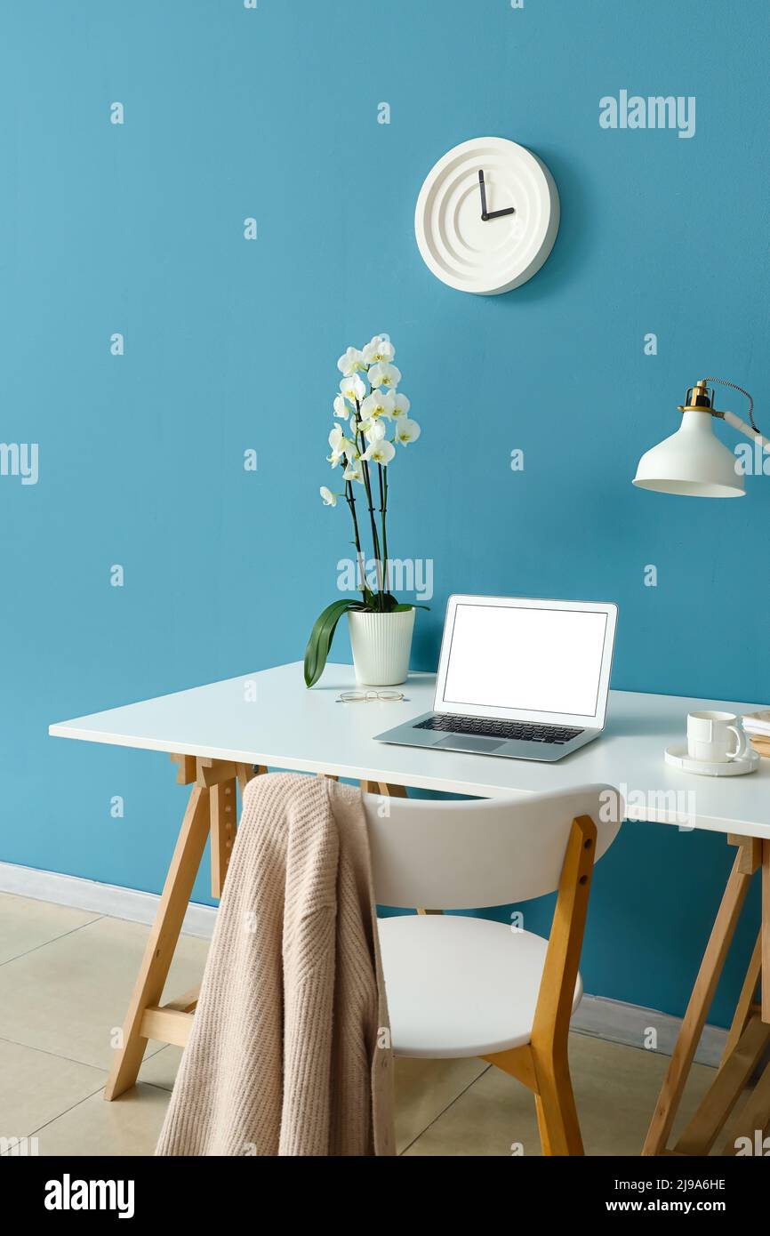 Workplace with modern laptop, orchid flower and cup near blue wall ...
