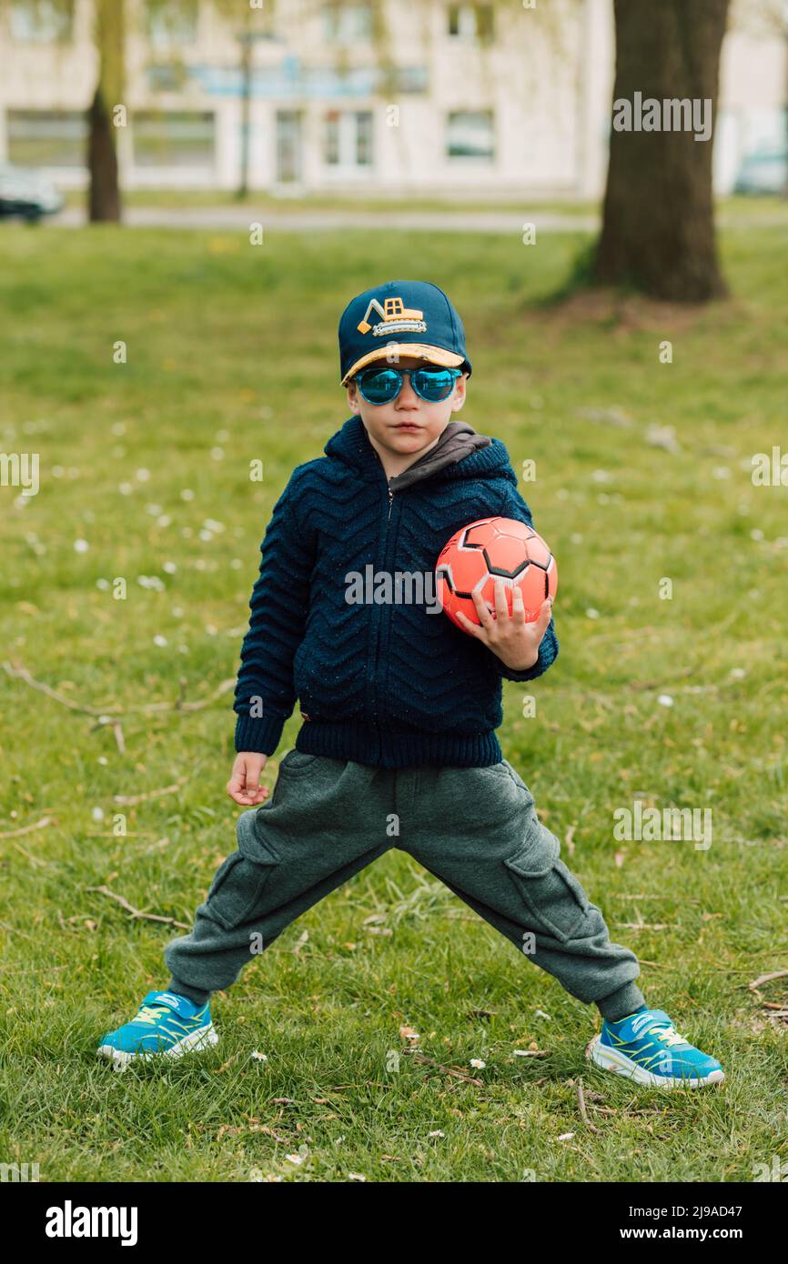A little boy stands in nature and holds an orange ball in his hand ...
