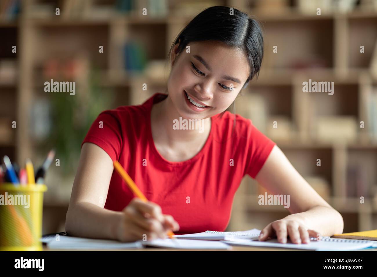 Cheerful pretty smart young chinese girl student making notes, homework ...