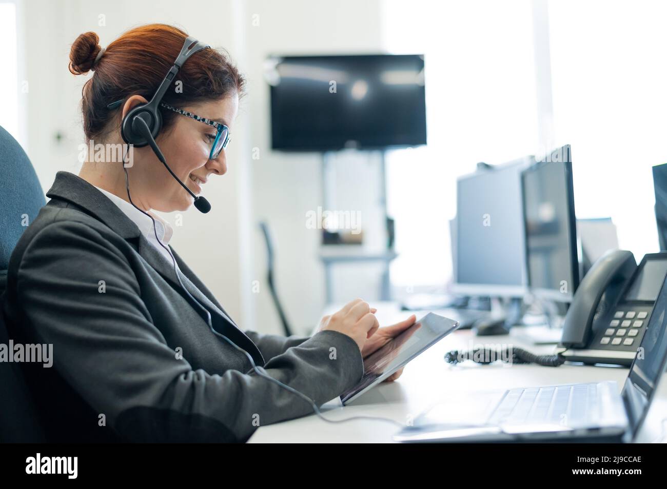 Woman in headset sits at her desk in the office and uses digital tablet ...