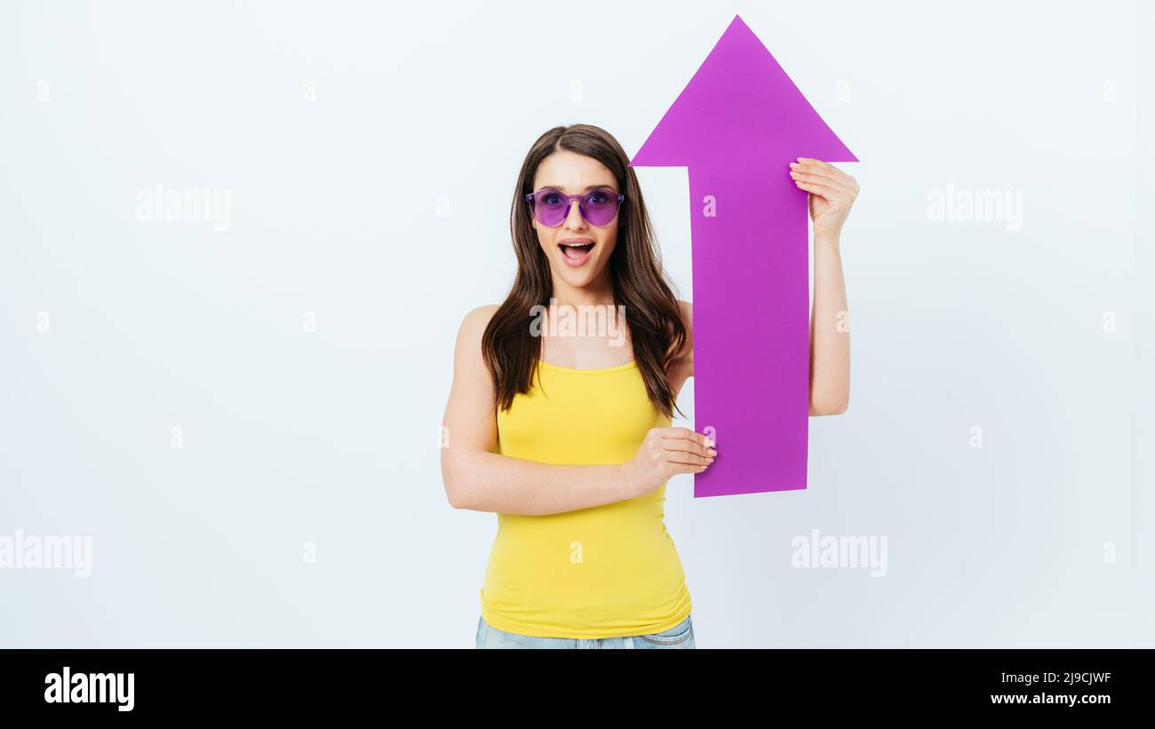 Surprised young woman wearing summer outfit holding big paper up arrow ...