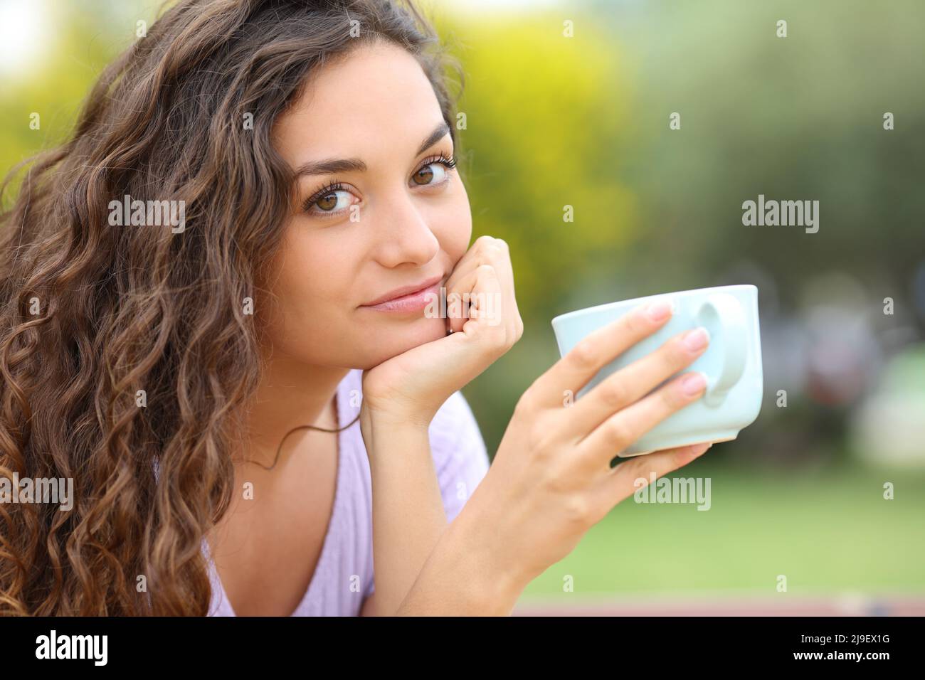 Confident woman looks at camera holding coffee mug sitting in a park ...