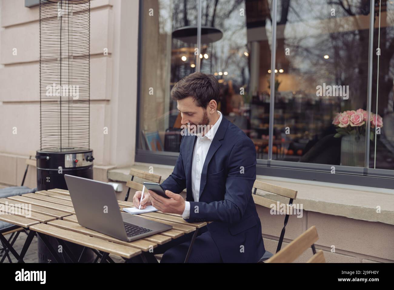 Smiled businessman working at laptop in cafe, holding phone and taking ...